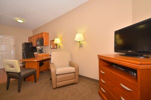 Quarto de hotel com cozinha compacta, mesa de jantar, sofá, televisão e armário de madeira.