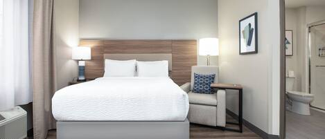 Quarto de hotel moderno com uma cama grande, abajures, uma cadeira e um banheiro visível através de uma porta aberta.
