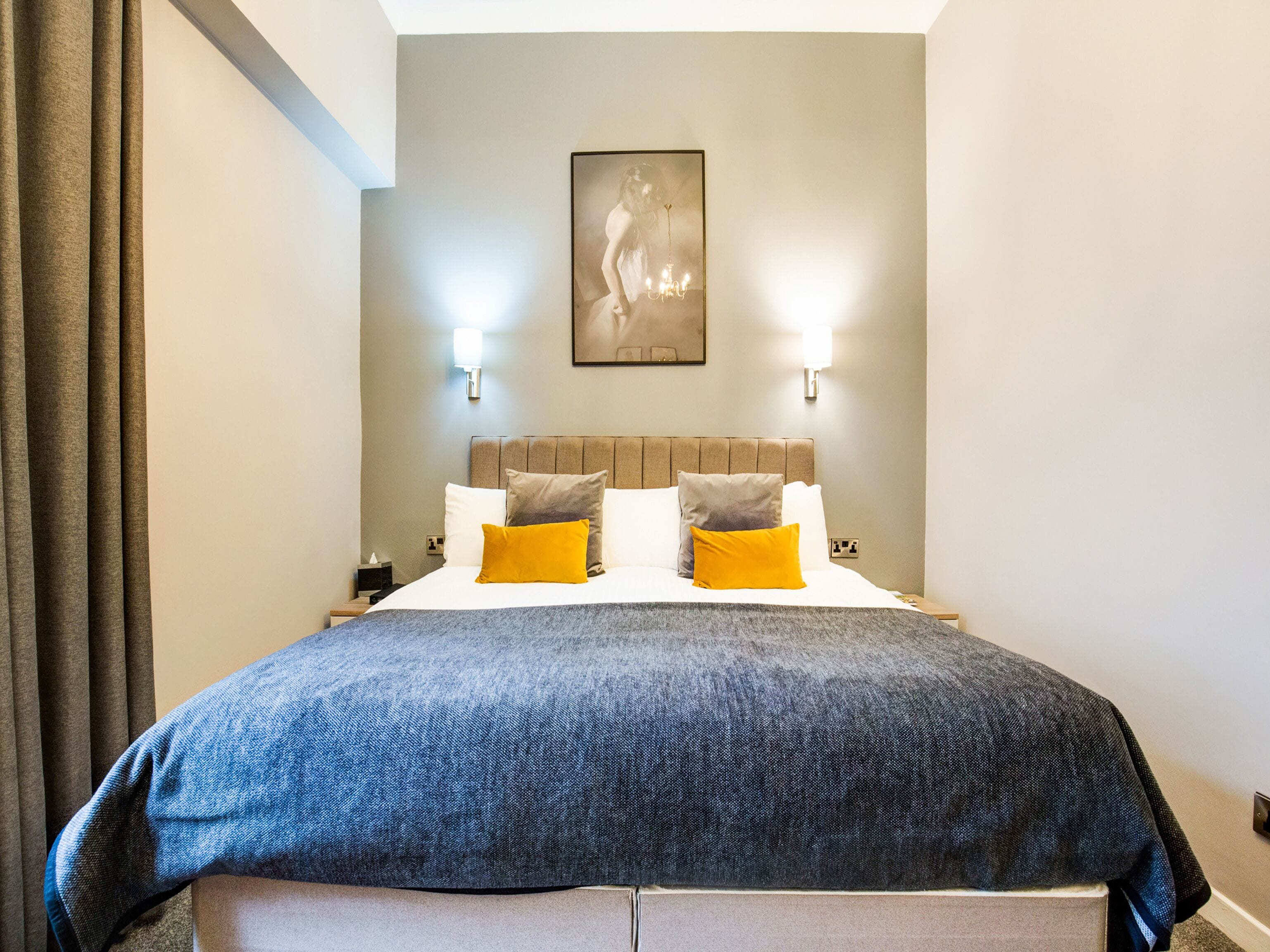 Cama bem arrumada com manta cinza, duas almofadas amarelas e cabeceira com luminária acima.