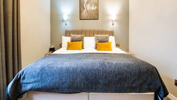 Cama bem arrumada com manta cinza, duas almofadas amarelas e cabeceira com luminária acima.