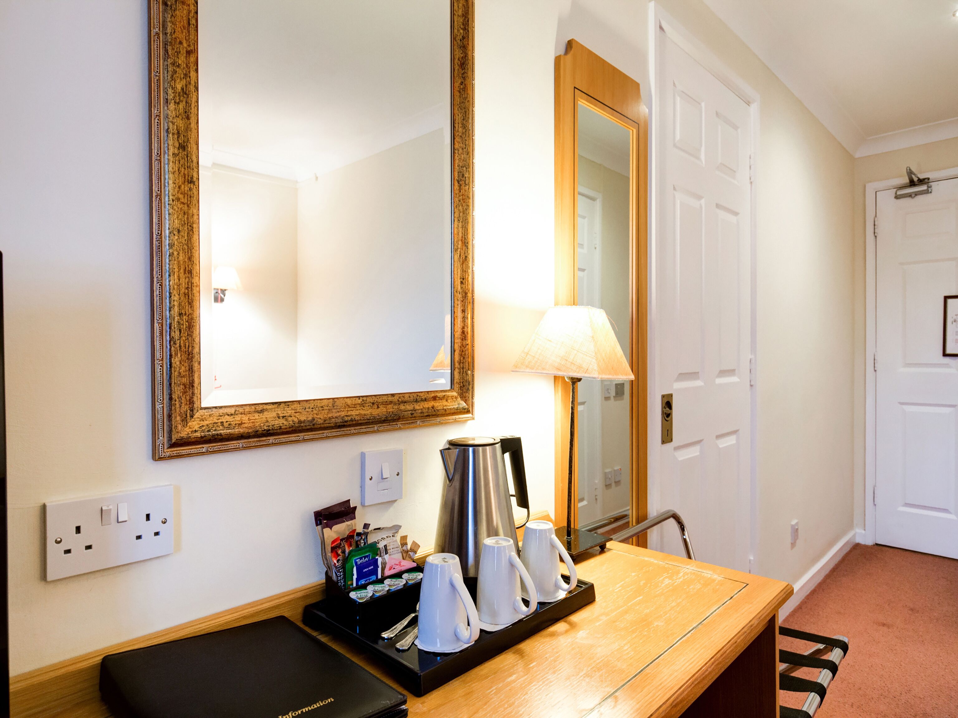 Quarto de hotel com escrivaninha de madeira, um espelho grande, uma luminária, um suporte com xícaras de café e um saco de artigos de higiene.