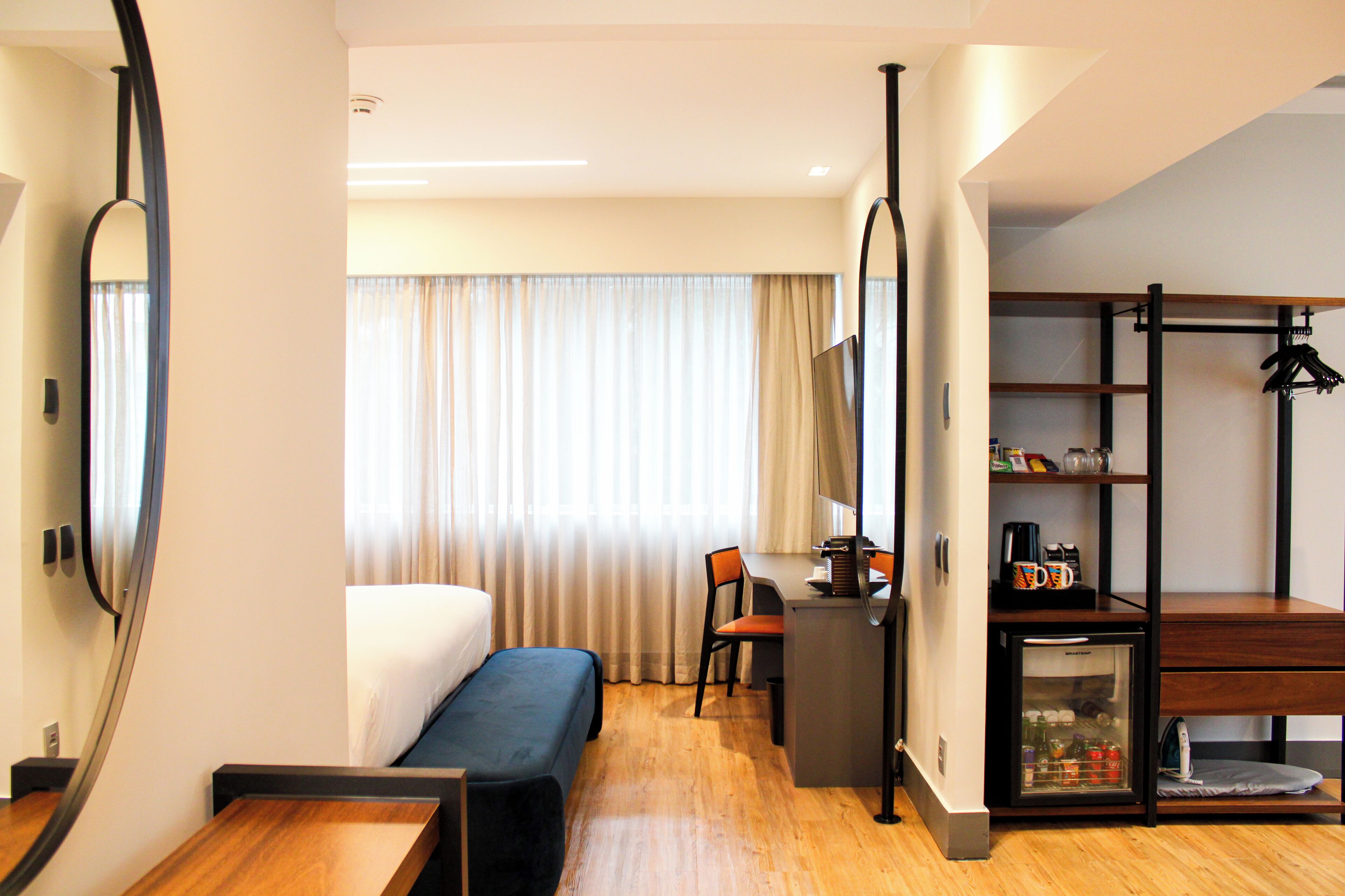 Quarto de hotel com uma cama, escrivaninha, cadeira, mini-frigorífico e guarda-roupa.