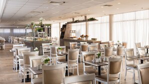 Uma ampla sala de jantar com várias mesas e cadeiras, um balcão de buffet e grandes janelas que oferecem iluminação natural.