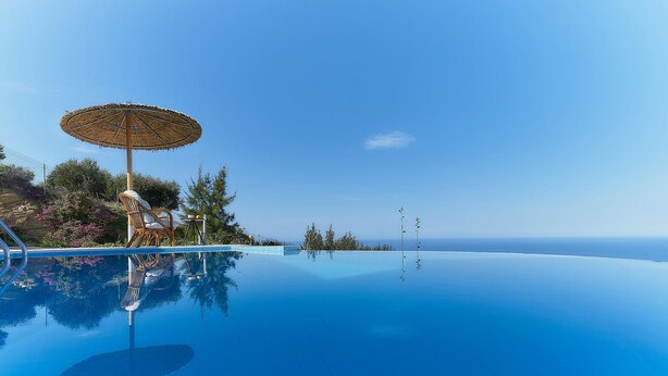 Uma piscina infinita azul cristalina com uma cadeira de vime e guarda-sol, rodeada por vegetação e com vista panorâmica.