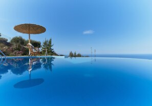 Uma piscina infinita azul cristalina com uma cadeira de vime e guarda-sol, rodeada por vegetação e com vista panorâmica.