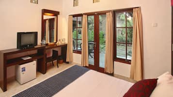 Quarto de hotel com cama, escrivaninha com televisão, pequeno frigorífico e vista para um jardim através de amplas janelas.