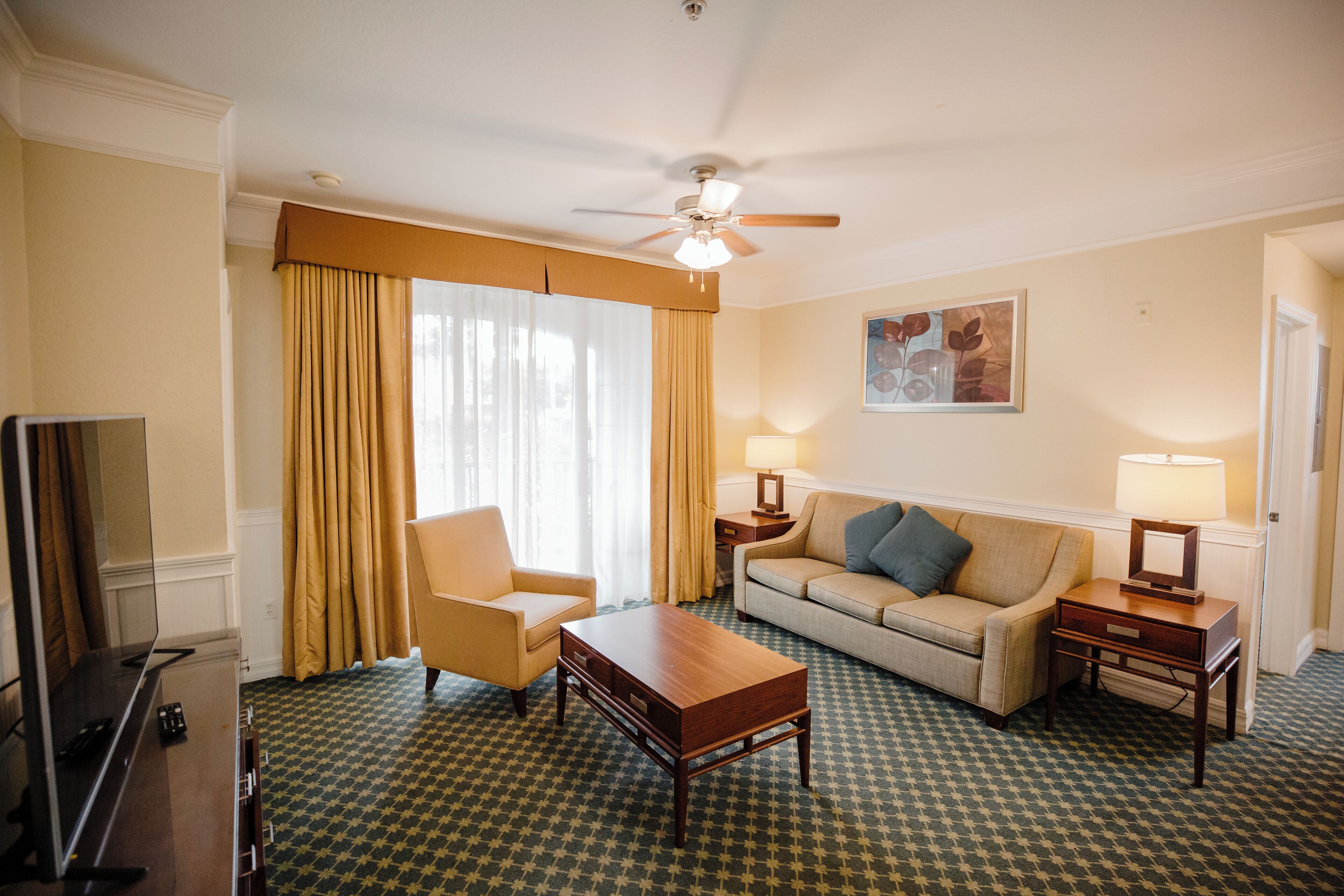 Quarto de hotel com sofá, poltrona, mesa de centro, TV, ventilador de teto e carpete estampado.