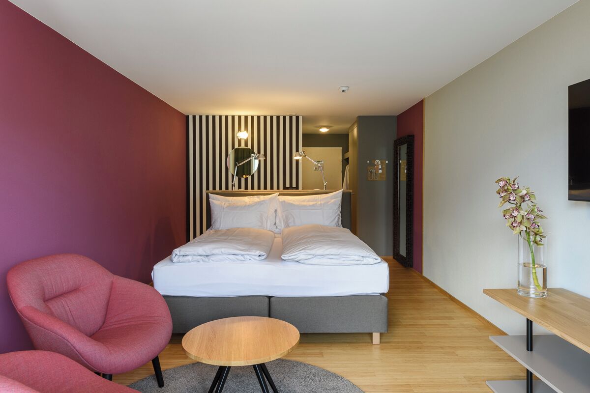 Quarto de hotel com cama, cadeira, mesa pequena, espelho e televisão.
