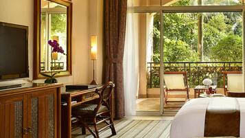 Quarto de hotel com uma cama grande, uma escrivaninha com cadeira, televisão, espelho e varanda com vista para vegetação.