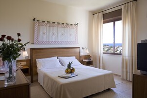 Cama bem arrumada com lençóis brancos, uma cesta de frutas na cama, um vaso com flores na mesa de cabeceira e vista para o mar pela janela.