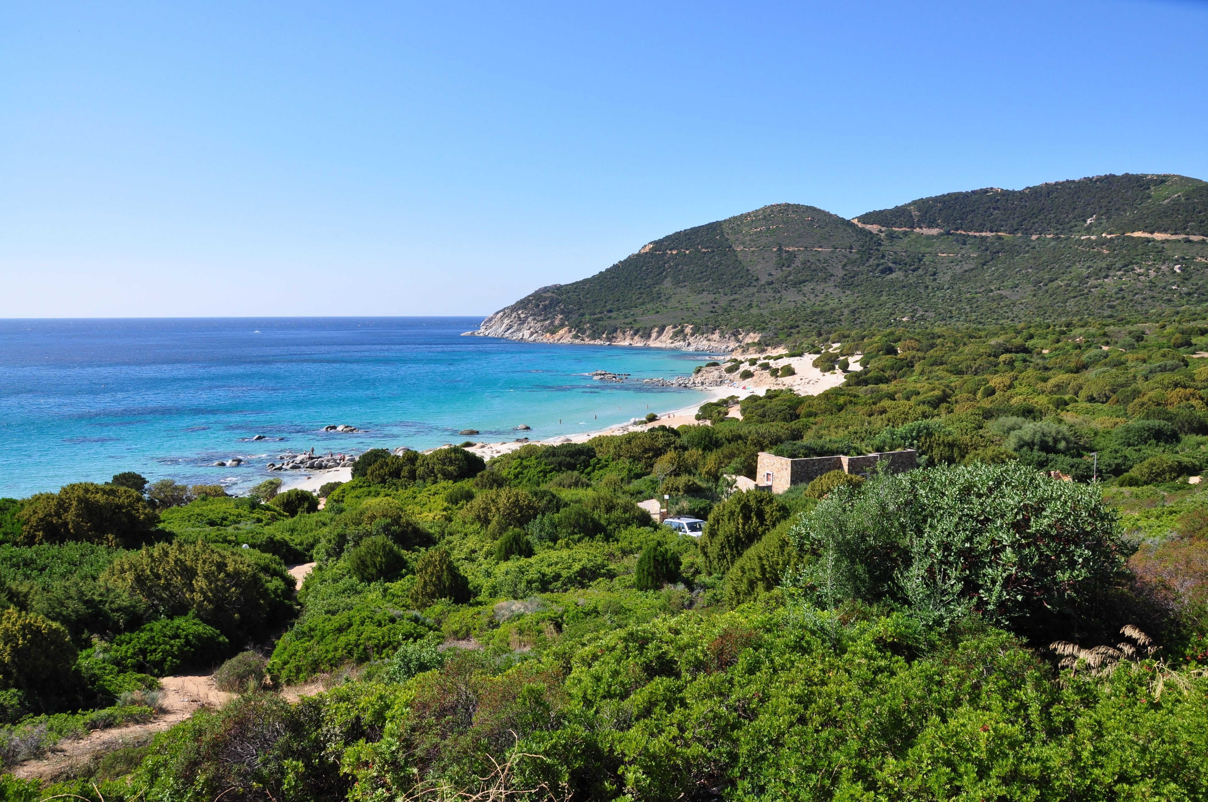Paisagem costeira com uma praia, vegetação densa e colinas ao fundo.