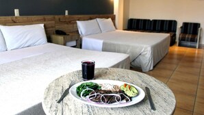 Quarto de hotel com duas camas, uma mesa de jantar com um prato de comida e um copo de vinho tinto, e um espaço de estar com cadeiras.