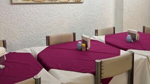 Uma sala de jantar com mesas cobertas por toalhas roxas, cadeiras brancas e um quadro pendurado na parede retratando vasos.