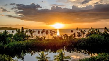 Um pôr do sol sobre águas calmas, com palmeiras e uma linha de costa distante.