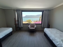 Quarto de hotel com duas camas, uma cadeira e uma janela grande com vista para o mar e montanhas.