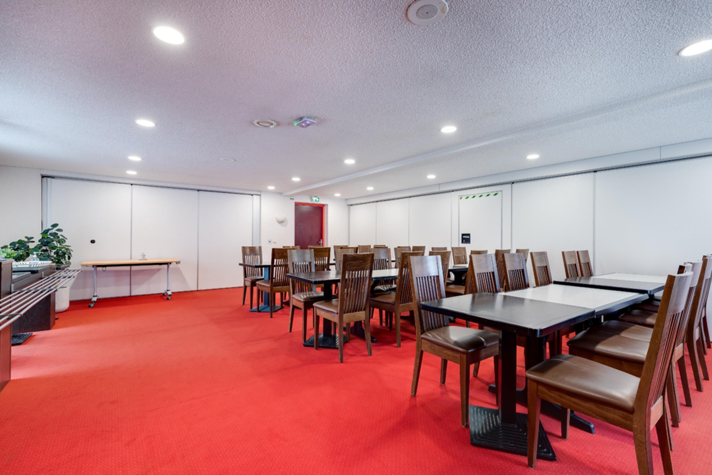 Uma sala de conferências com várias mesas e cadeiras, piso de tapete vermelho, paredes brancas e luminárias no teto.