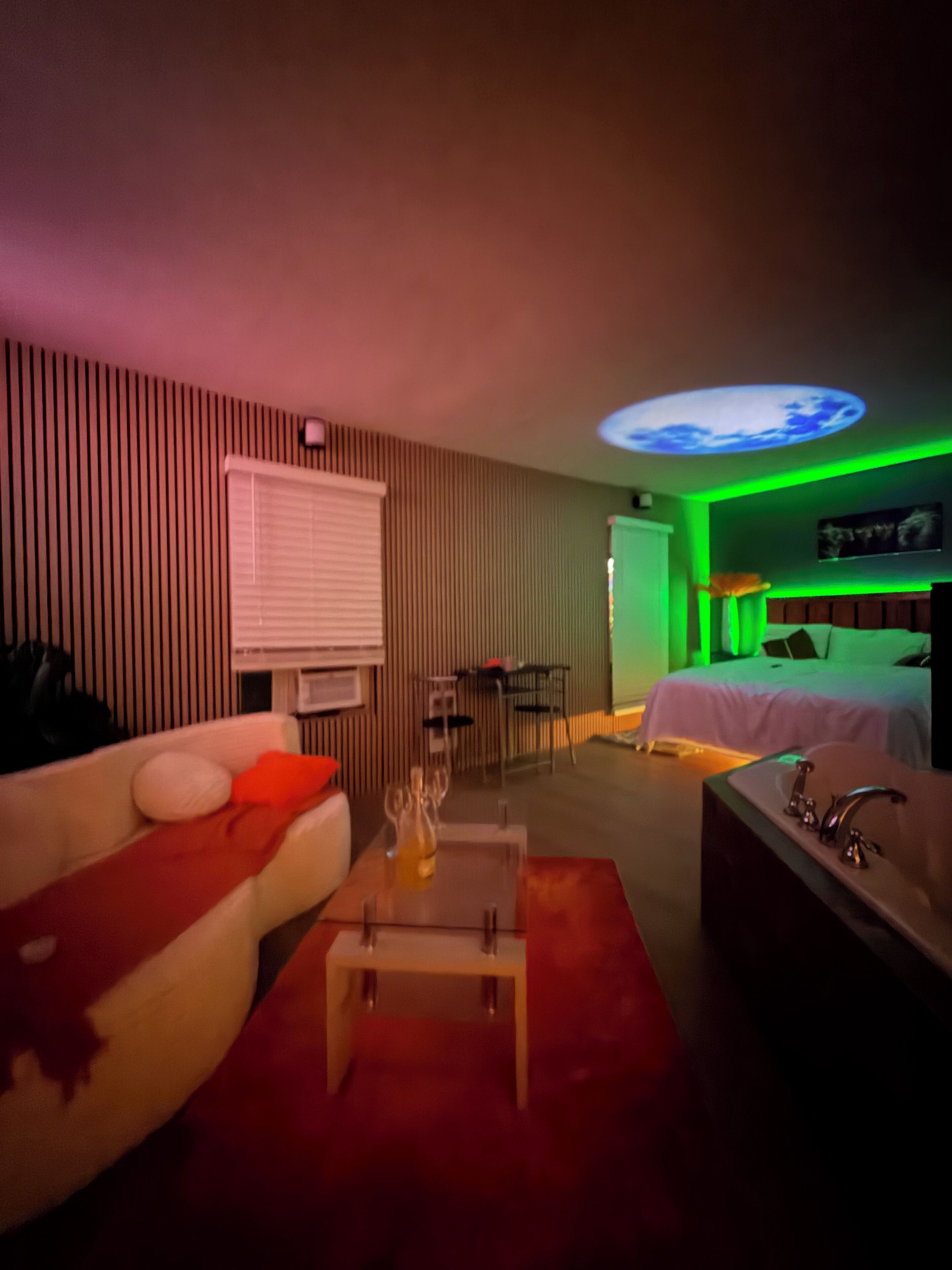 Quarto de hotel moderno com sofá, mesa de centro, cama, banheiro e luminária no teto.