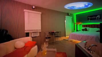 Quarto de hotel moderno com sofá, mesa de centro, cama, banheiro e luminária no teto.