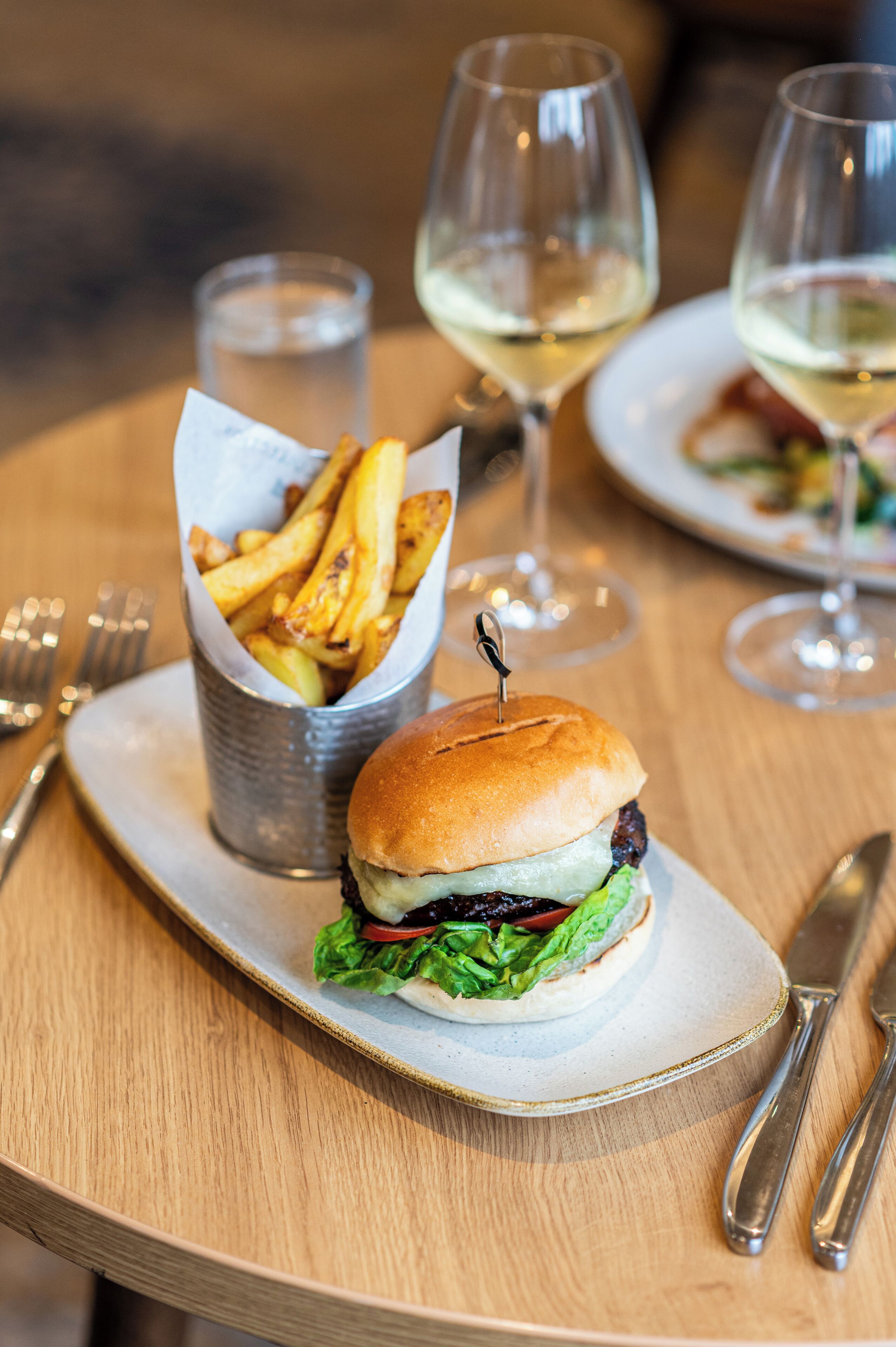Uma refeição com hambúrguer, batatas fritas e copos de vinho sobre uma mesa de madeira.