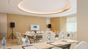 Sala de conferência com uma mesa comprida, cadeiras brancas, garrafas de água, tela de projetor e alto-falantes.