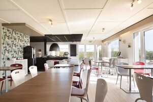 Uma área de jantar moderna com uma mesa de madeira comprida e cadeiras, cozinha ao fundo e grandes janelas que proporcionam luz natural.