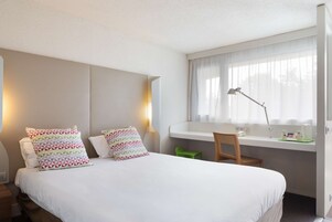 Quarto de hotel com cama, escrivaninha, cadeira e janela com cortinas.