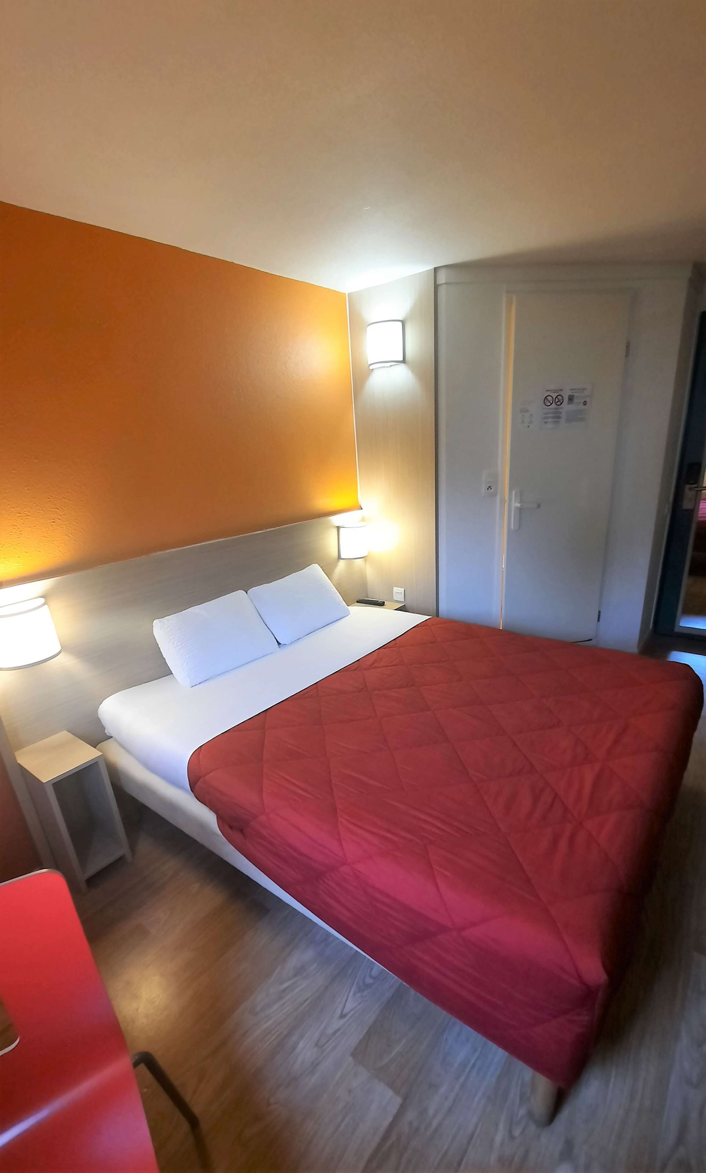 Quarto de hotel com uma cama, mesa de cabeceira, luminária, guarda-roupa e uma cadeira vermelha.