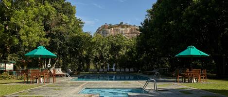 Piscina com espreguiçadeiras e guarda-sóis, cercada por árvores e vista para uma montanha.