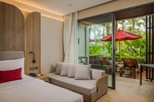 Quarto de hotel moderno com cama, sofá, uma pequena mesa e porta de vidro que dá para uma varanda com mobiliário e vegetação.