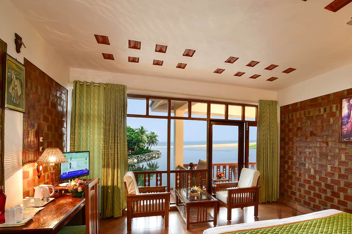 Quarto com uma grande janela que oferece vista para o mar, uma escrivaninha de madeira com um computador, uma cama e um espaço de estar com cadeiras e uma mesa.
