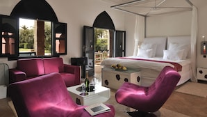Quarto com cama grande, área de estar com sofá e poltronas, pequena mesa com garrafa e copos, e vista para o exterior através de janelas em arco.