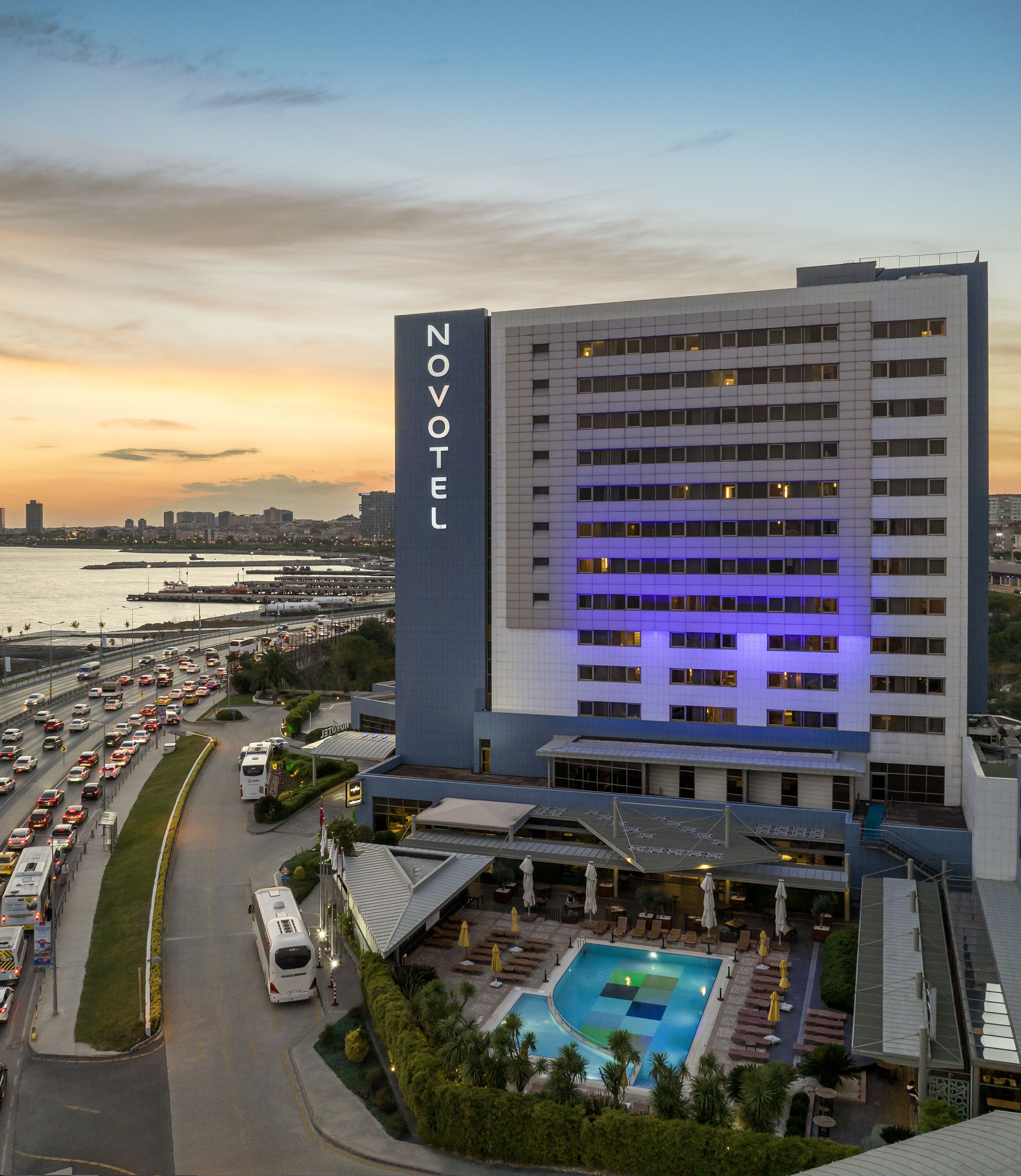 Um hotel com uma placa chamativa da 'NOVOVOTEL', uma piscina no terraço e vista para a paisagem urbana e o mar.
