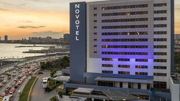 Um hotel com uma placa chamativa da 'NOVOVOTEL', uma piscina no terraço e vista para a paisagem urbana e o mar.
