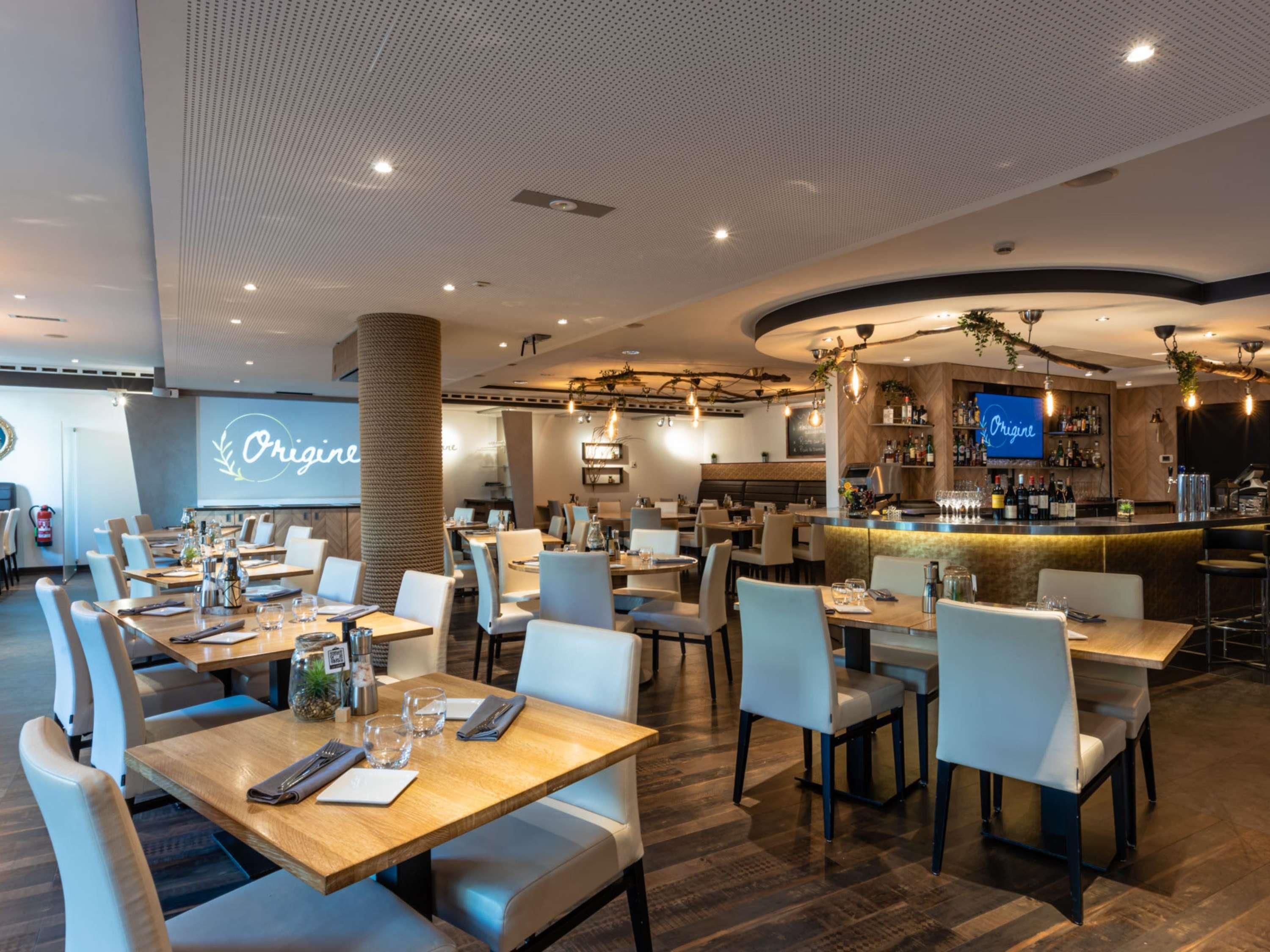Um restaurante moderno com área de bar, mesas de madeira e cadeiras brancas.