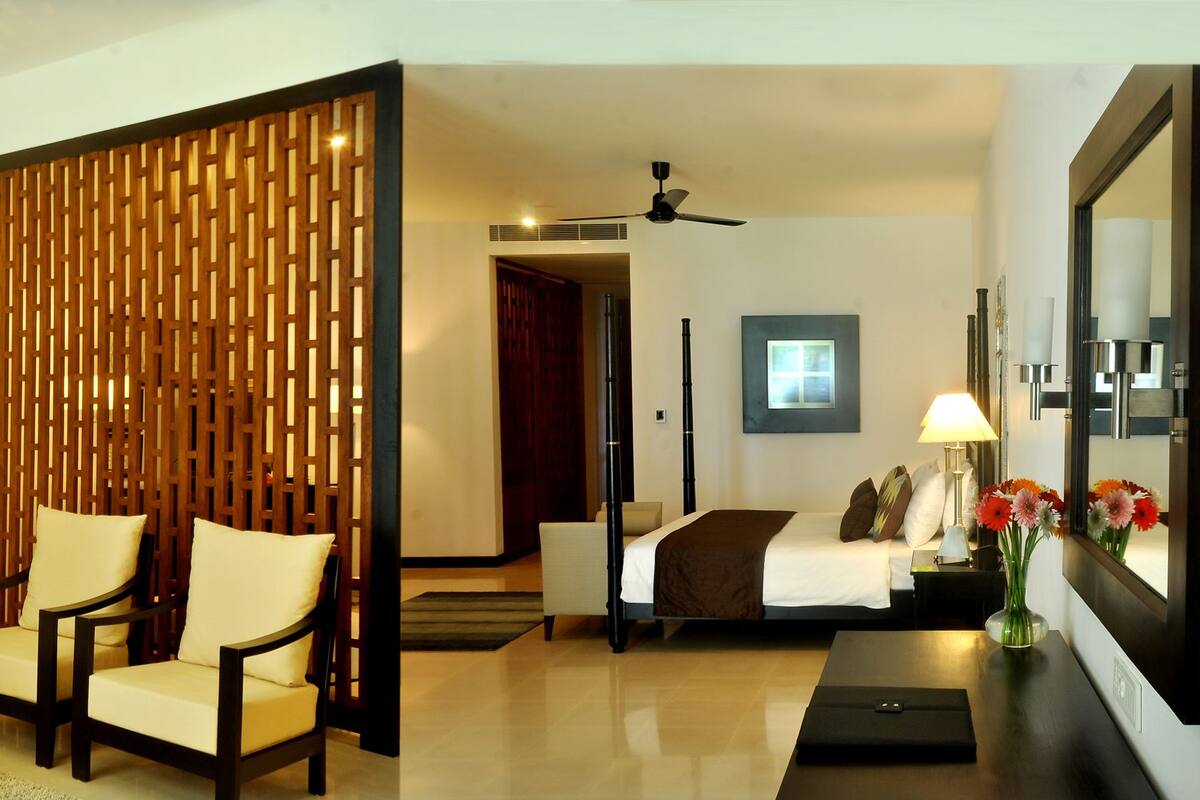Quarto de hotel com sofá, cadeiras, cama, mesa de cabeceira, abajur, espelho e um quadro.