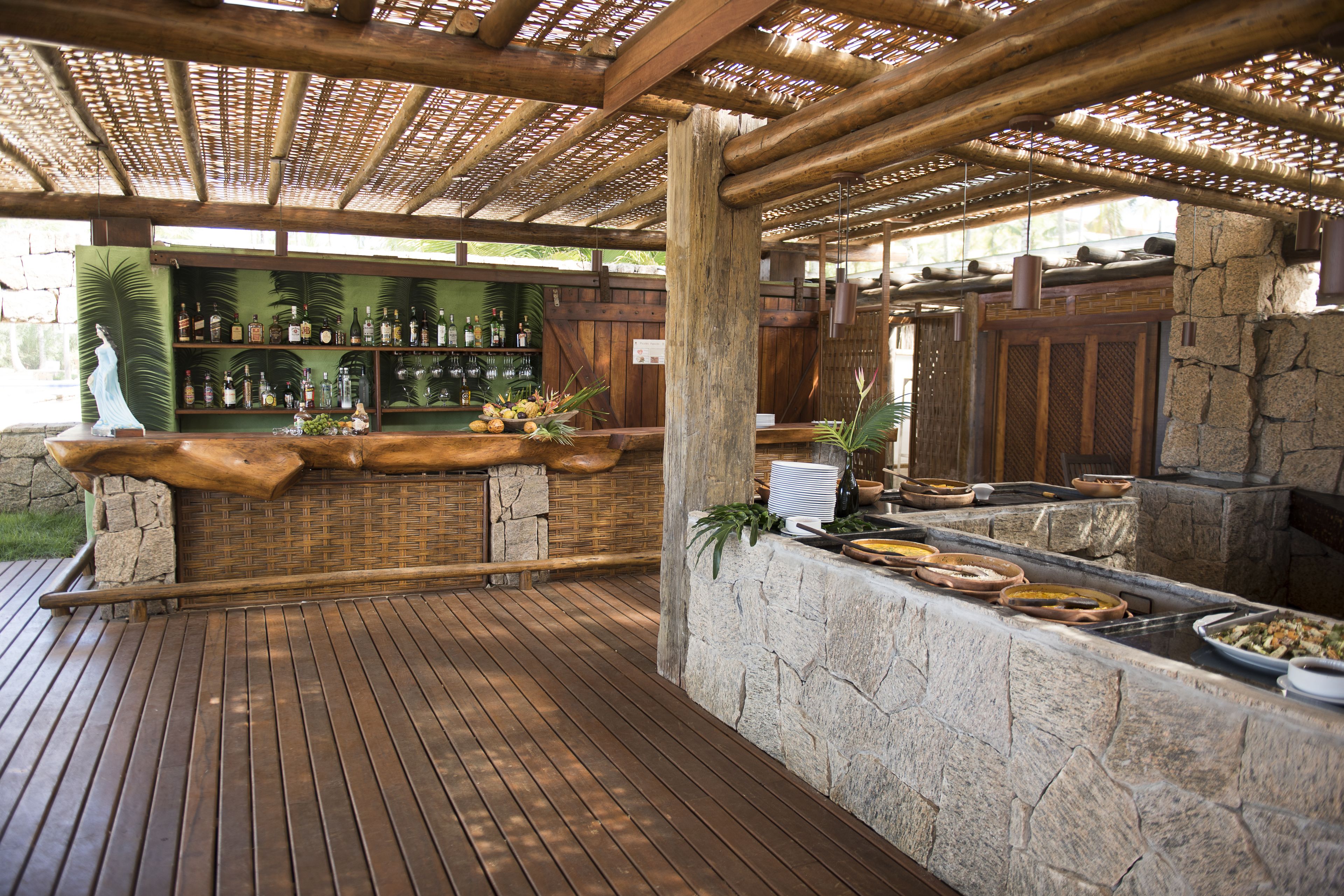 Uma área de bar ao ar livre coberta, com piso de madeira, paredes de pedra e teto de bambu.