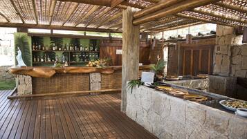 Uma área de bar ao ar livre coberta, com piso de madeira, paredes de pedra e teto de bambu.