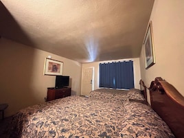 Quarto de hotel com cama, televisão num penteadeira, porta e janela com cortinas.