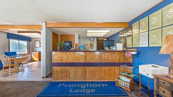 Uma área de recepção com uma mesa de madeira, um tapete azul com a inscrição 'Pronghorn Lodge', uma parede com prêmios emoldurados e vista para um quarto com cama e janela.