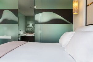 Quarto de hotel moderno com cama, banheiro com espelho e luminária na parede.
