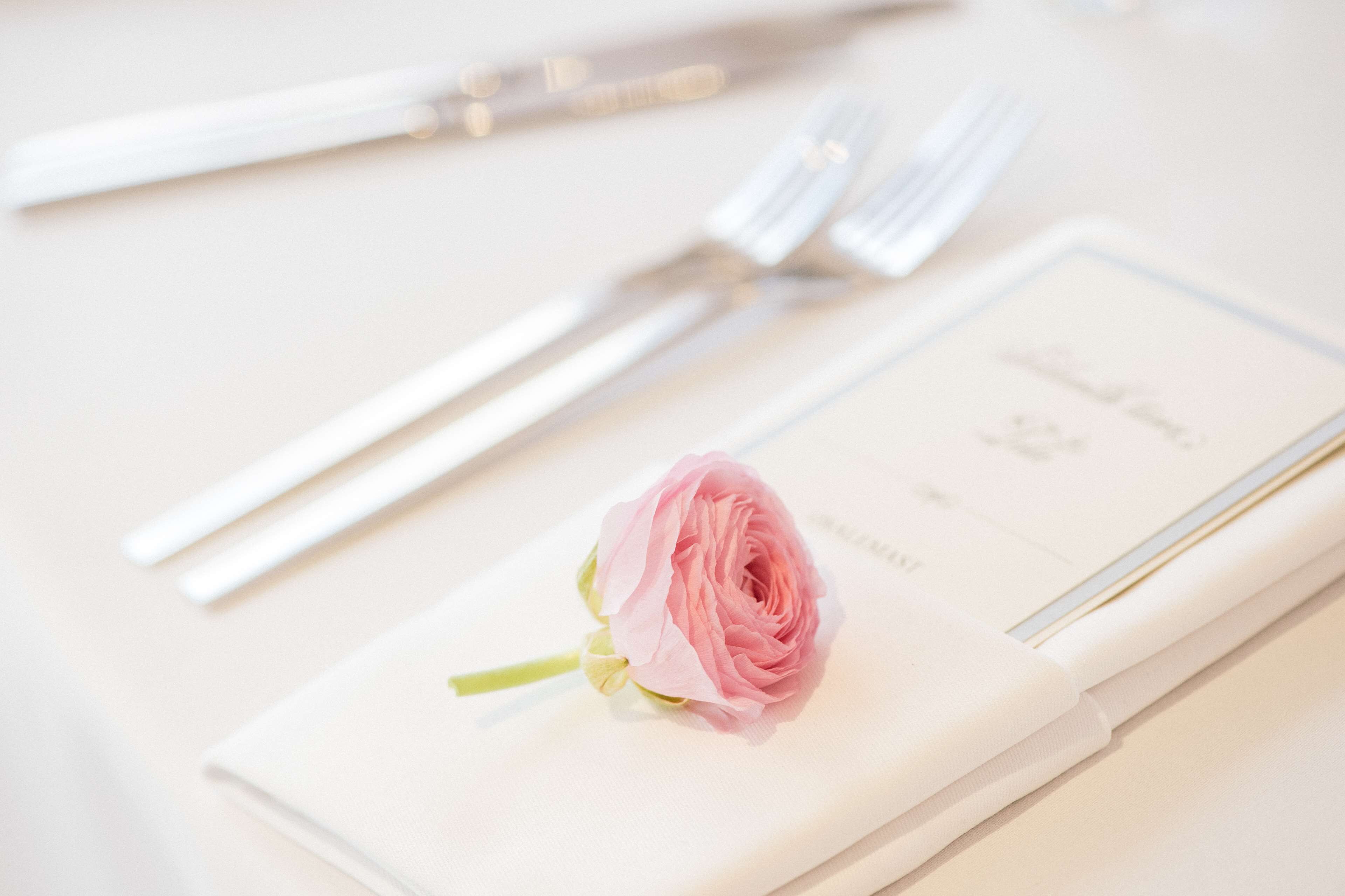 Uma mesa bem posta, com um guardanapo dobrado, uma rosa cor-de-rosa e talheres de prata.