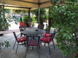 Pátio externo com mesa redonda e quatro cadeiras, rodeado por vasos de plantas e um toldo amarelo.