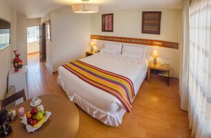 Quarto de hotel com uma cama grande, mesas de cabeceira com luminárias, uma pequena mesa com fruta e uma chávena, e vista para o corredor.