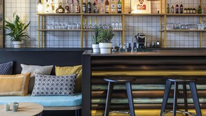 Uma área de bar com prateleiras de garrafas e copos, um sofá com almofadas estampadas e um pendente.