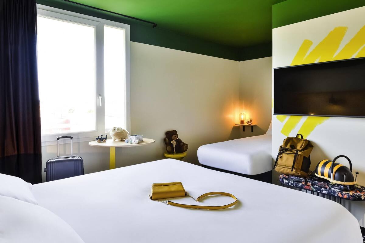 Quarto de hotel com cama, uma mesinha com abajur, televisão e mala.
