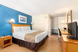 Quarto de hotel com cama, mesas de cabeceira, escrivaninha com computador, televisão e espelho.