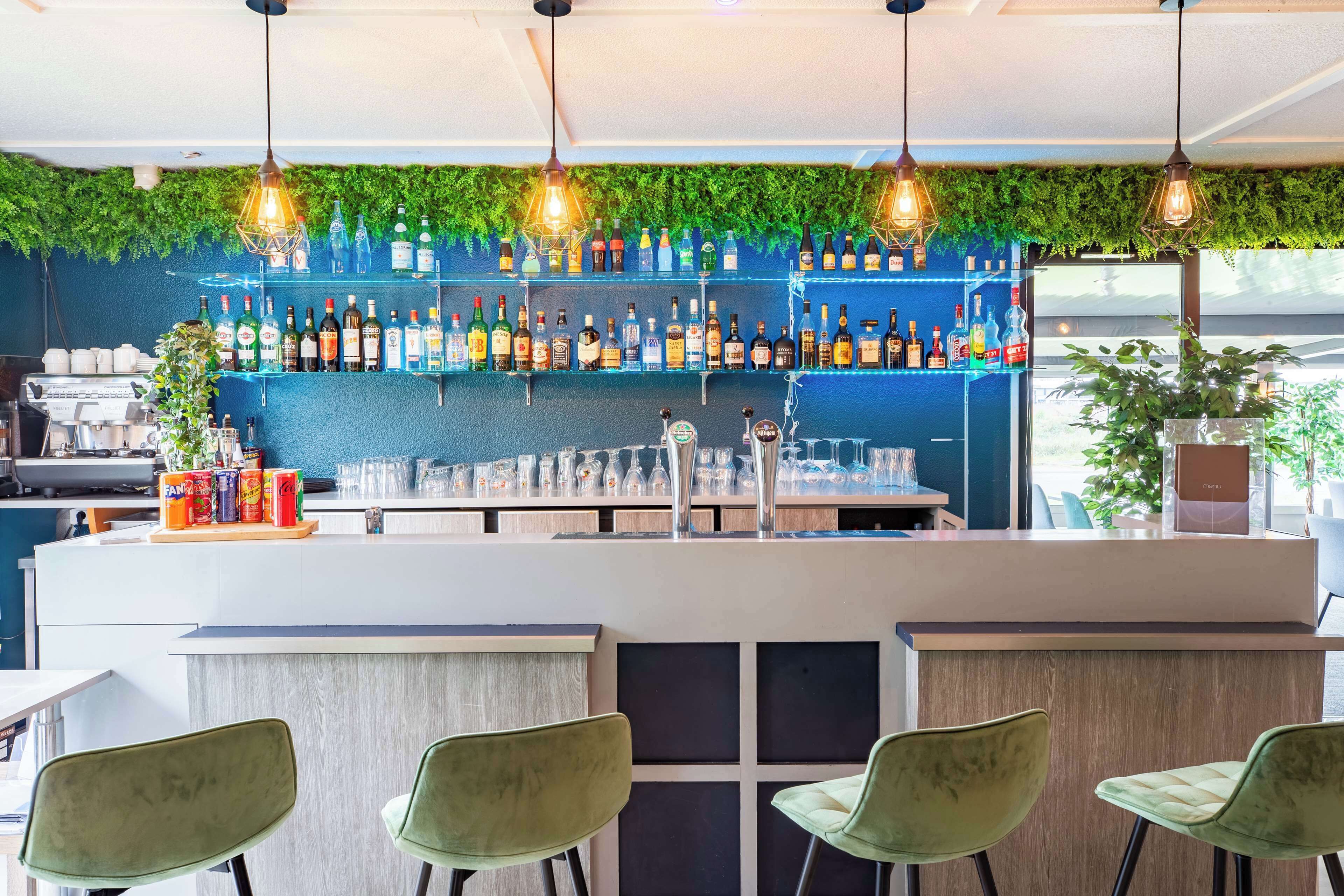 Uma área de bar com parede azul, plantas verdes e luminárias penduradas. Há banquetas e prateleiras com garrafas.