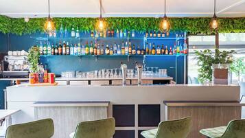 Uma área de bar com parede azul, plantas verdes e luminárias penduradas. Há banquetas e prateleiras com garrafas.