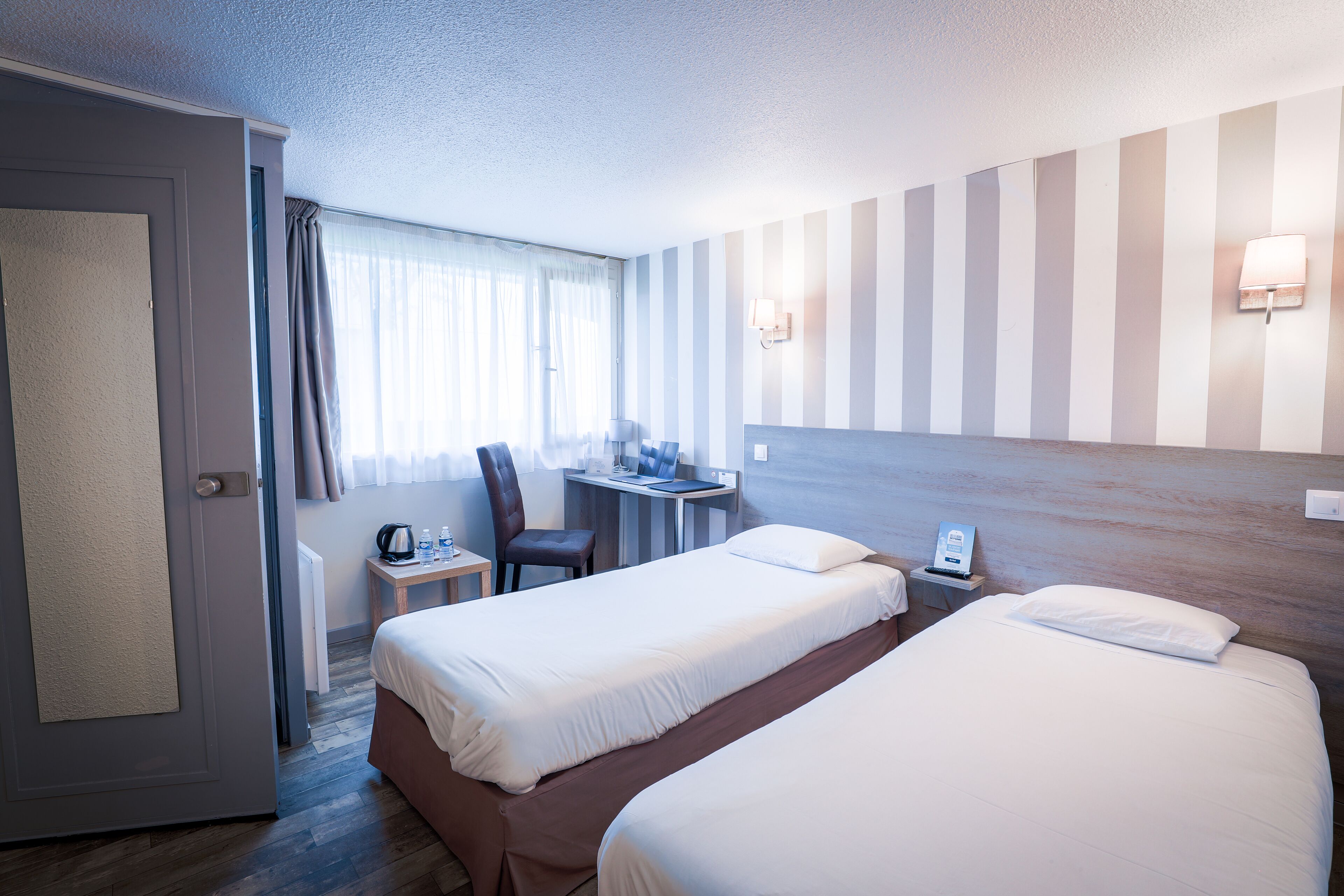 Quarto de hotel com duas camas, uma escrivaninha, uma cadeira e um abajur.