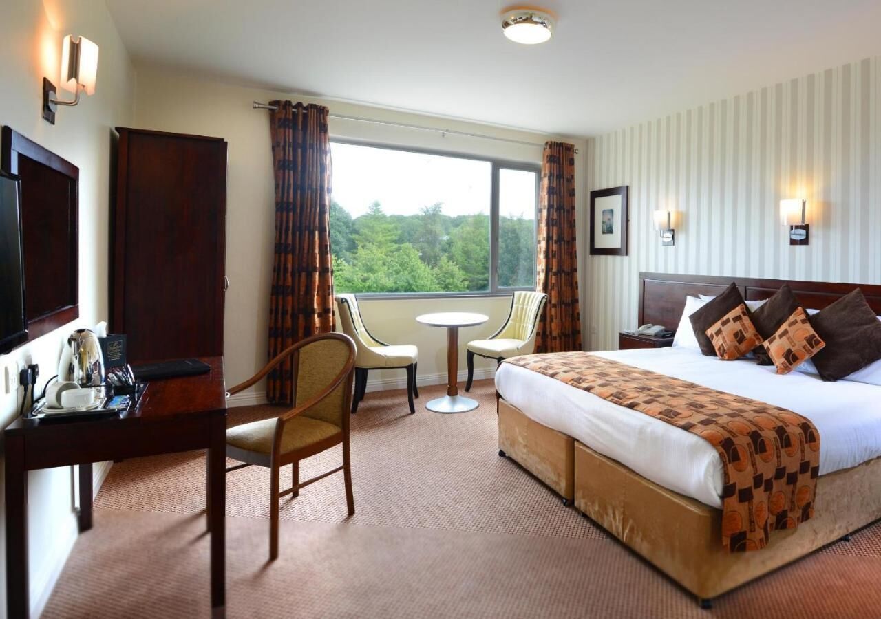 Quarto de hotel com cama, escrivaninha, duas cadeiras, uma mesa pequena e janela com cortinas.
